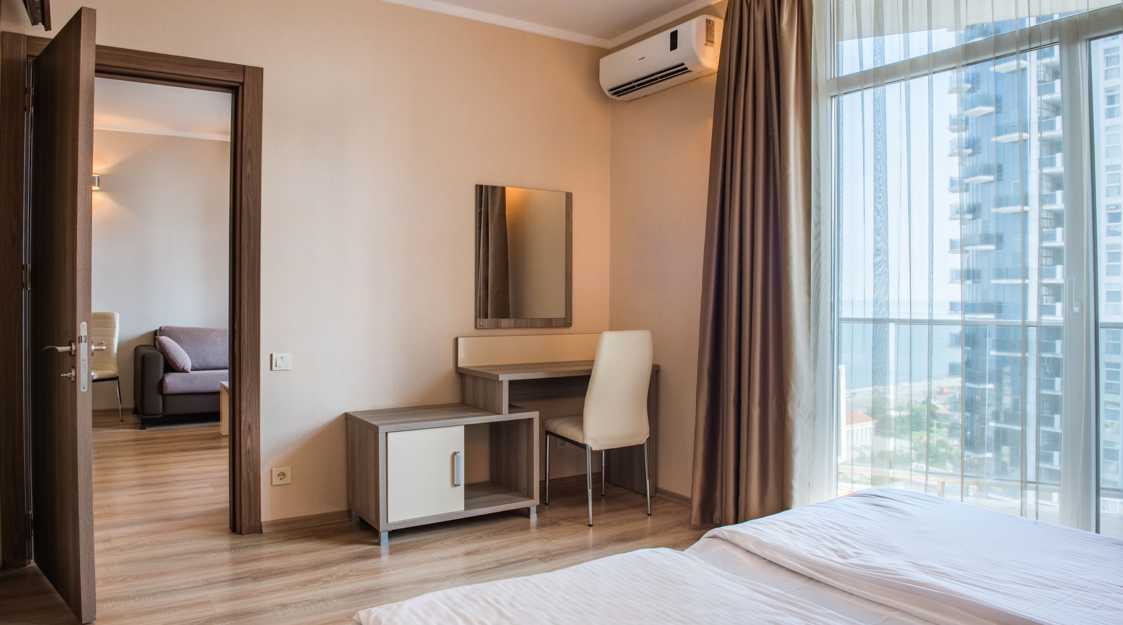 Quarto de hotel com cama, escrivaninha, cadeira, guarda-roupa, espelho e uma janela grande com vista para edifícios.