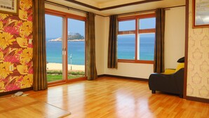 Quarto espaçoso com piso de madeira, amplas janelas com cortinas, uma cadeira e vista para o mar.