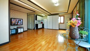Uma sala de estar ampla com piso de madeira, uma mesa de jantar de vidro com cadeiras, uma cozinha ao fundo e um vaso decorativo com flores na mesa.