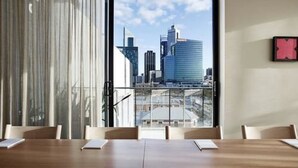 Uma sala de conferência com uma mesa grande e cadeiras, vista para a cidade através de janelas amplas, e um sinal vermelho em formato de 'X' na parede.