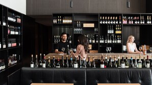 Um bar com várias prateleiras de garrafas de vinho e copos, um balcão e assentos de couro preto.