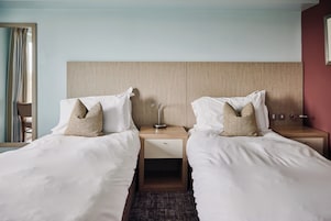 Quarto de hotel com duas camas de solteiro, uma mesa de cabeceira com abajur e cabeceira de madeira.