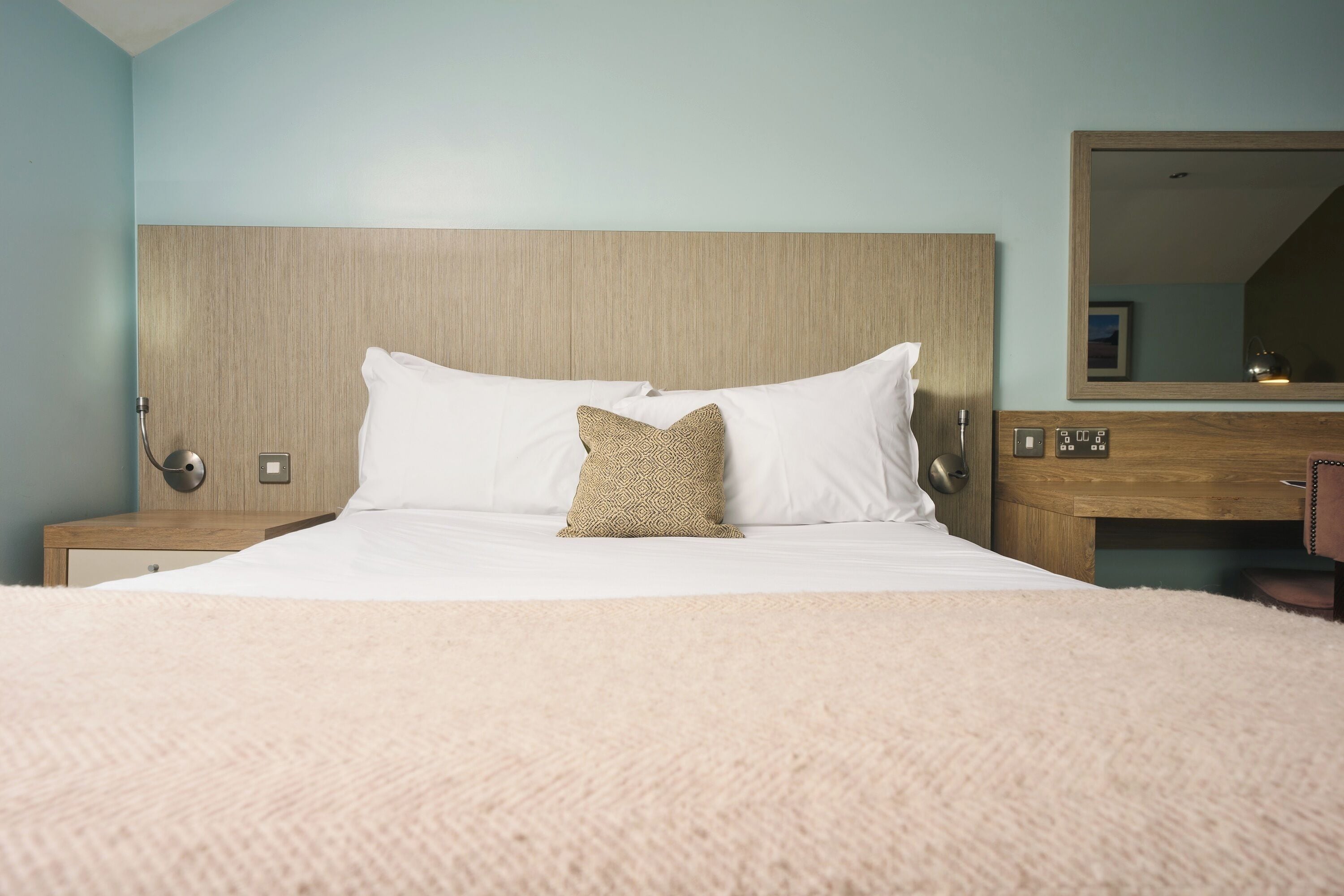 Quarto de hotel com uma cama grande, cabeceira de madeira, mesa de cabeceira, espelho e cômoda com espelho.