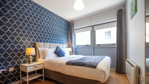 Quarto bem arrumado com cama, mesas de cabeceira, luminária e janela com cortinas.