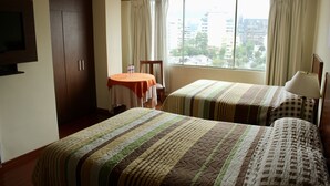 Quarto de hotel com duas camas, uma mesinha com cadeira, televisão e janela com cortinas.