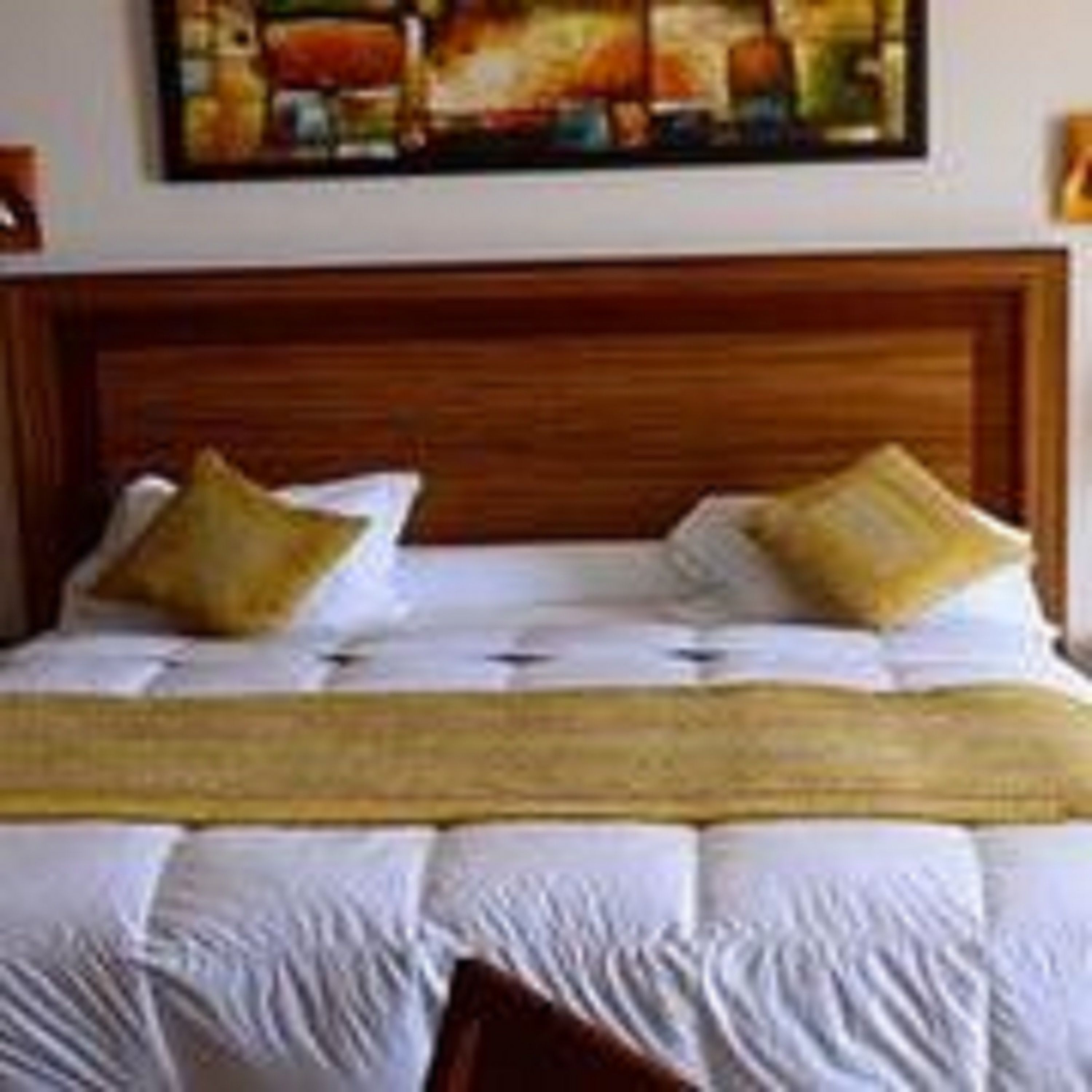 Cama bem arrumada com roupa de cama branca e duas almofadas, cabeceira de madeira e um quadro na parede acima.