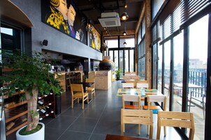 Interior de restaurante com mesas e cadeiras de madeira, uma planta grande em vaso e um mural na parede.