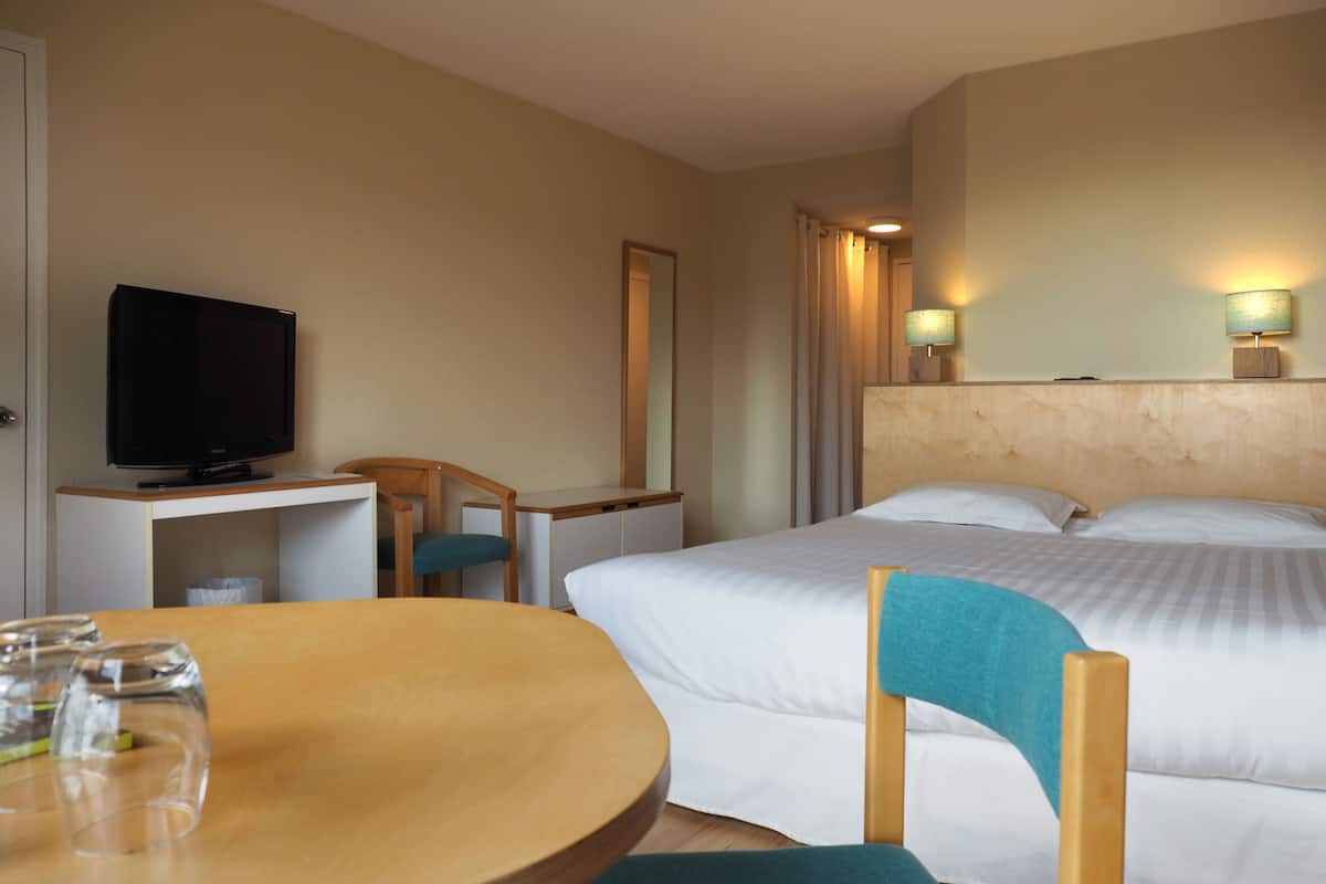 Quarto de hotel com cama, escrivaninha com televisão, cadeira e mesa com copos.