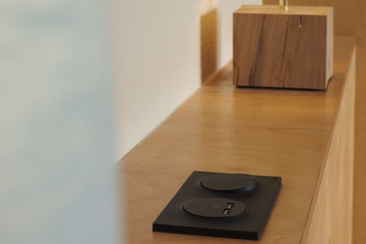Uma mesa de cabeceira de madeira com uma luminária moderna e um pequeno dispositivo eletrônico.