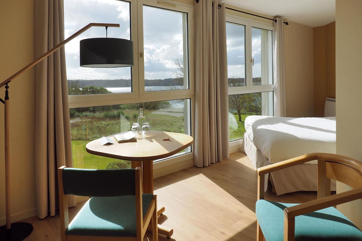 Quarto de hotel com cama, escrivaninha com cadeira, mesa com cadeira e grandes janelas com vista para o exterior.