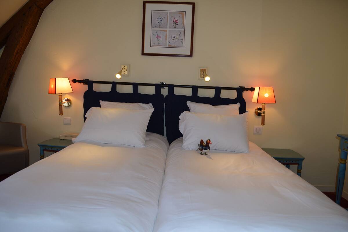 Uma cama de casal com lençóis brancos, duas mesas de cabeceira com abajures e uma obra de arte emoldurada sobre a cama.