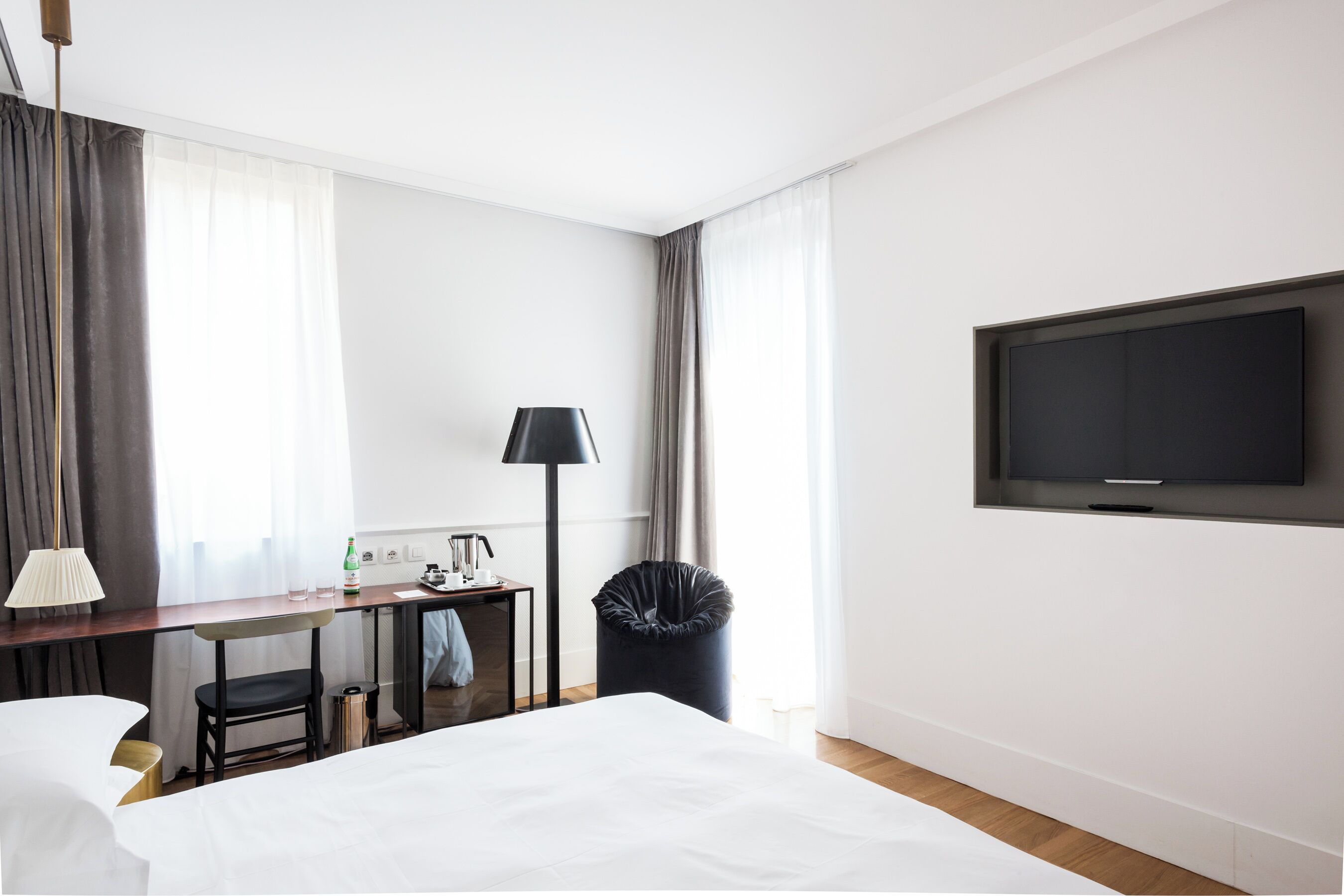Quarto de hotel com cama, escrivaninha com abajur, televisão na parede e lixeira.