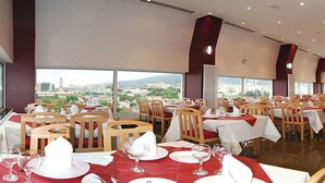 Restaurante com várias mesas preparadas para refeições, grandes janelas com vista para a cidade e design de interiores moderno.