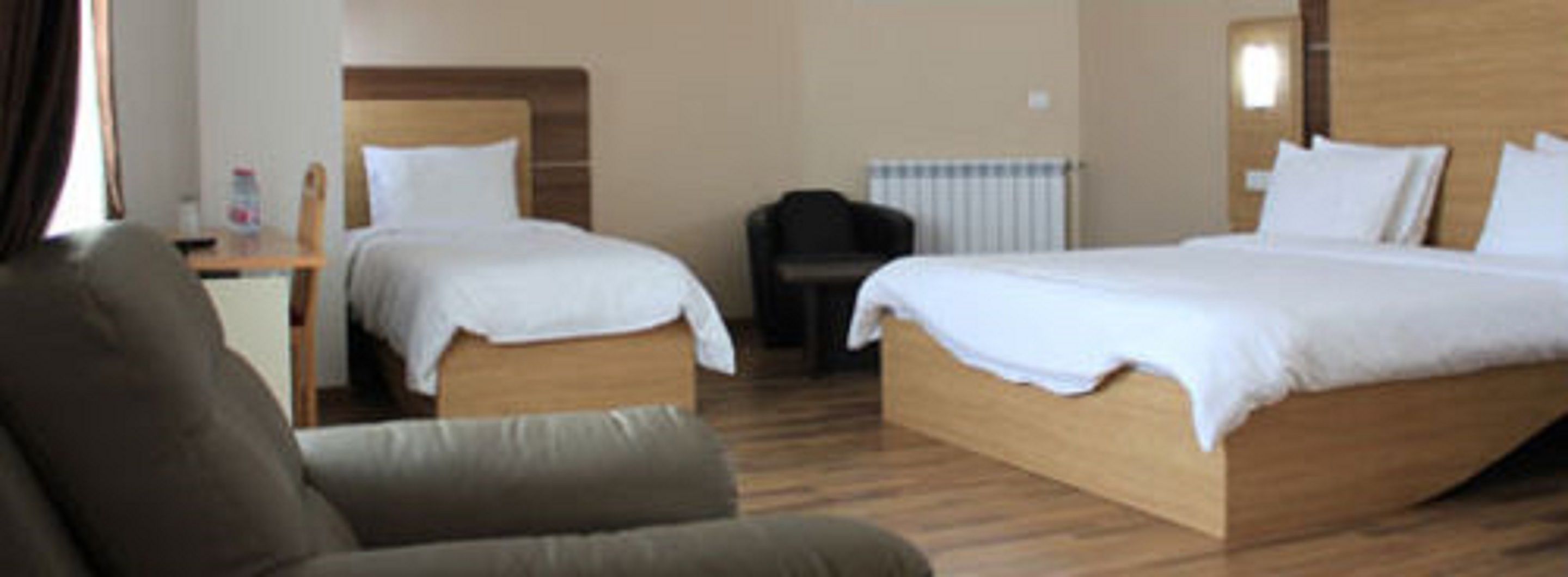 Quarto de hotel com duas camas de solteiro, cabeceira de madeira, uma secretária pequena com cadeira e radiador.