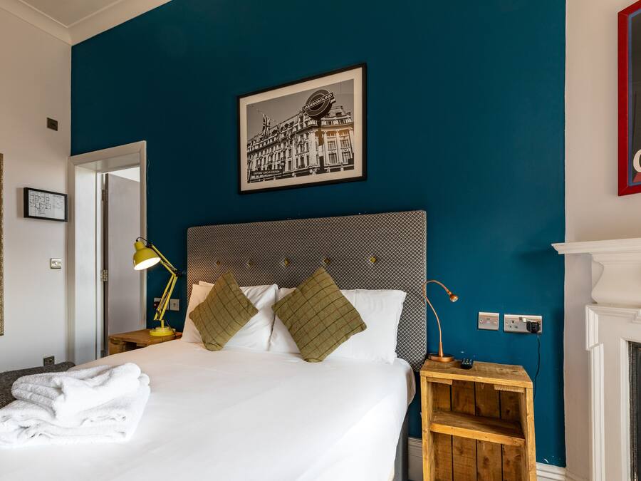 Cama bem arrumada com lençóis brancos e duas almofadas verdes, uma moldura com fotografia de um edifício, um espelho com moldura dourada e uma mesa de cabeceira com abajur.