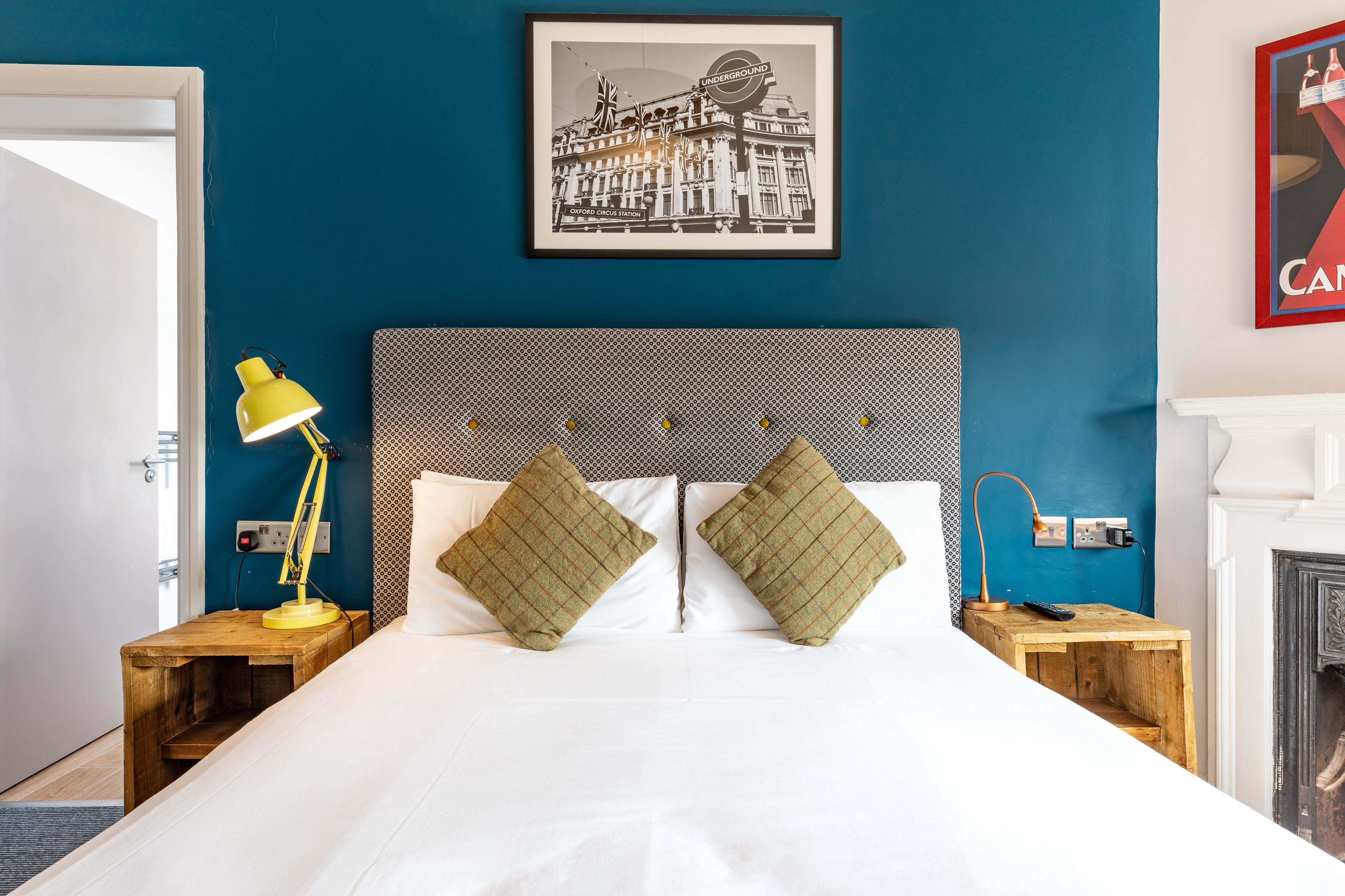 Cama bem arrumada com lençóis brancos e duas almofadas verdes, um quadro emoldurado acima dela e uma pequena mesa de cabeceira de madeira com um abajur amarelo ao lado.