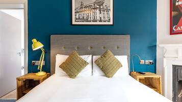 Cama bem arrumada com lençóis brancos e duas almofadas verdes, um quadro emoldurado acima dela e uma pequena mesa de cabeceira de madeira com um abajur amarelo ao lado.