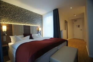 Quarto de hotel com uma cama grande, cabeceira com design estampado, mesa de cabeceira com abajur e porta que dá para outro cômodo.
