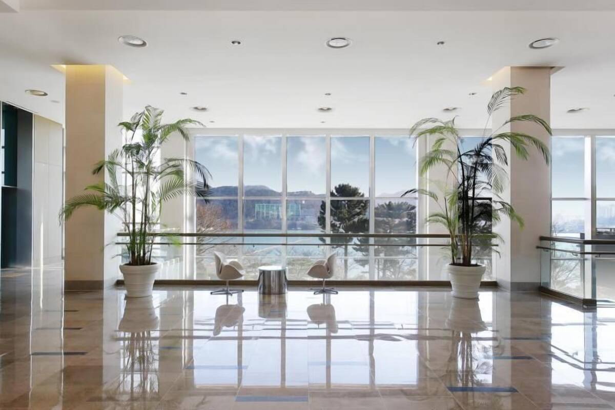 Um hall amplo com piso refletivo, grandes janelas com vista para a cidade e plantas em vasos.