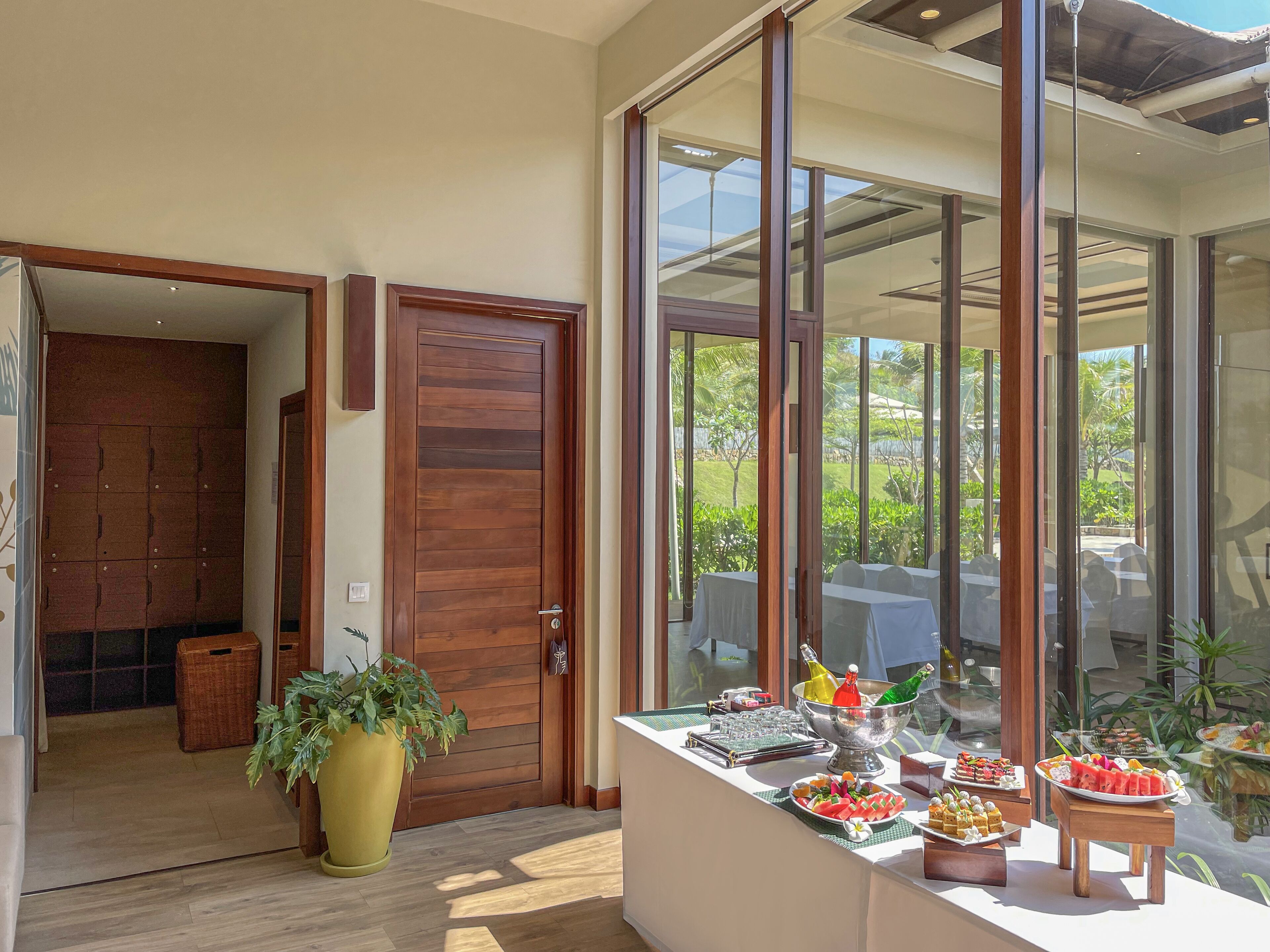 Quarto com porta de madeira, uma grande planta em vaso e uma mesa de buffet com diversos alimentos.