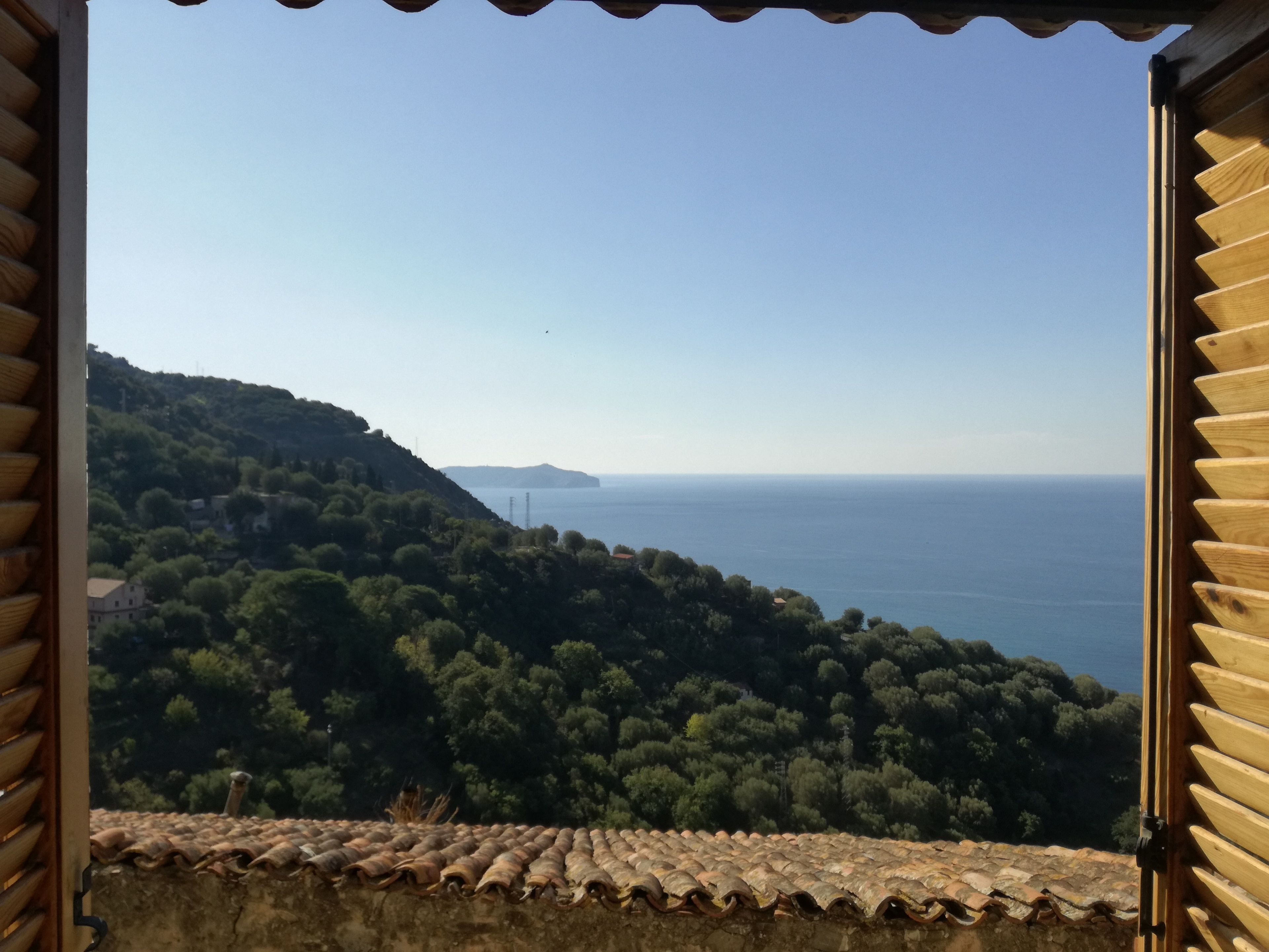 Vista de um quarto com telhado telhado e persianas de madeira, com vista para uma paisagem costeira com vegetação densa e o mar.