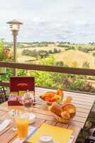 Uma mesa de café da manhã com um cesto de croissants, uma fruteira, um bule de chá e um copo de suco em uma varanda com vista para o campo.