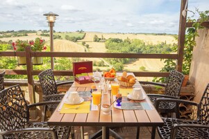 Mesa de madeira posta com itens de café da manhã, incluindo croissants e suco de laranja, em uma varanda com uma vista panorâmica de campos.