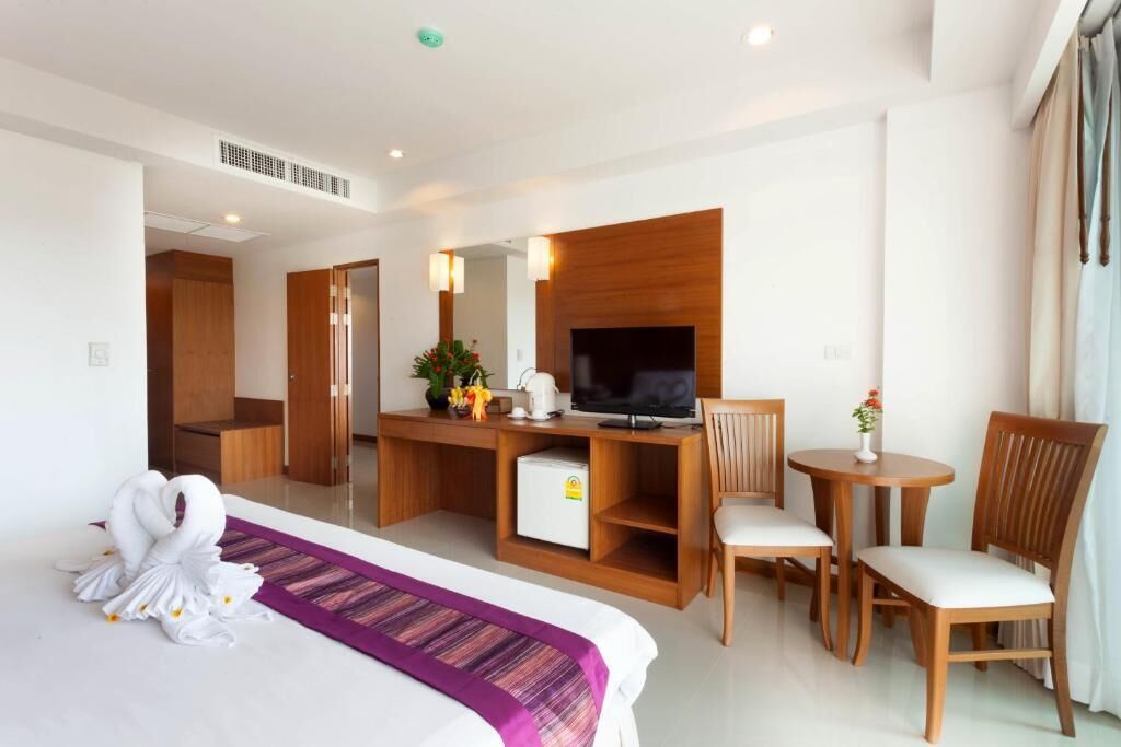 Quarto de hotel com cama, escrivaninha, televisão, mini-geladeira e duas cadeiras.