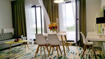 Quarto de hotel com área de refeições, sofá, uma pequena mesa com frutas e vista através de grandes janelas.