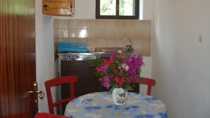 Uma cozinha pequena com janela, pia e fogão. Há uma mesa com toalha de mesa florida e um vaso com flores.