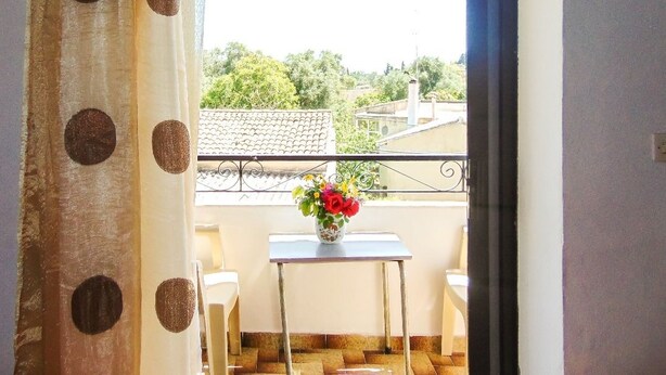 Uma varanda com mesa e um vaso de flores, uma cadeira branca e vista para edifícios e árvores.
