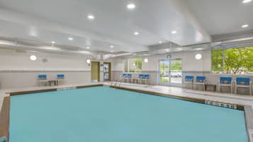 Uma grande piscina interior com água cristalina, rodeada por cadeiras azuis e um chão em mosaico.