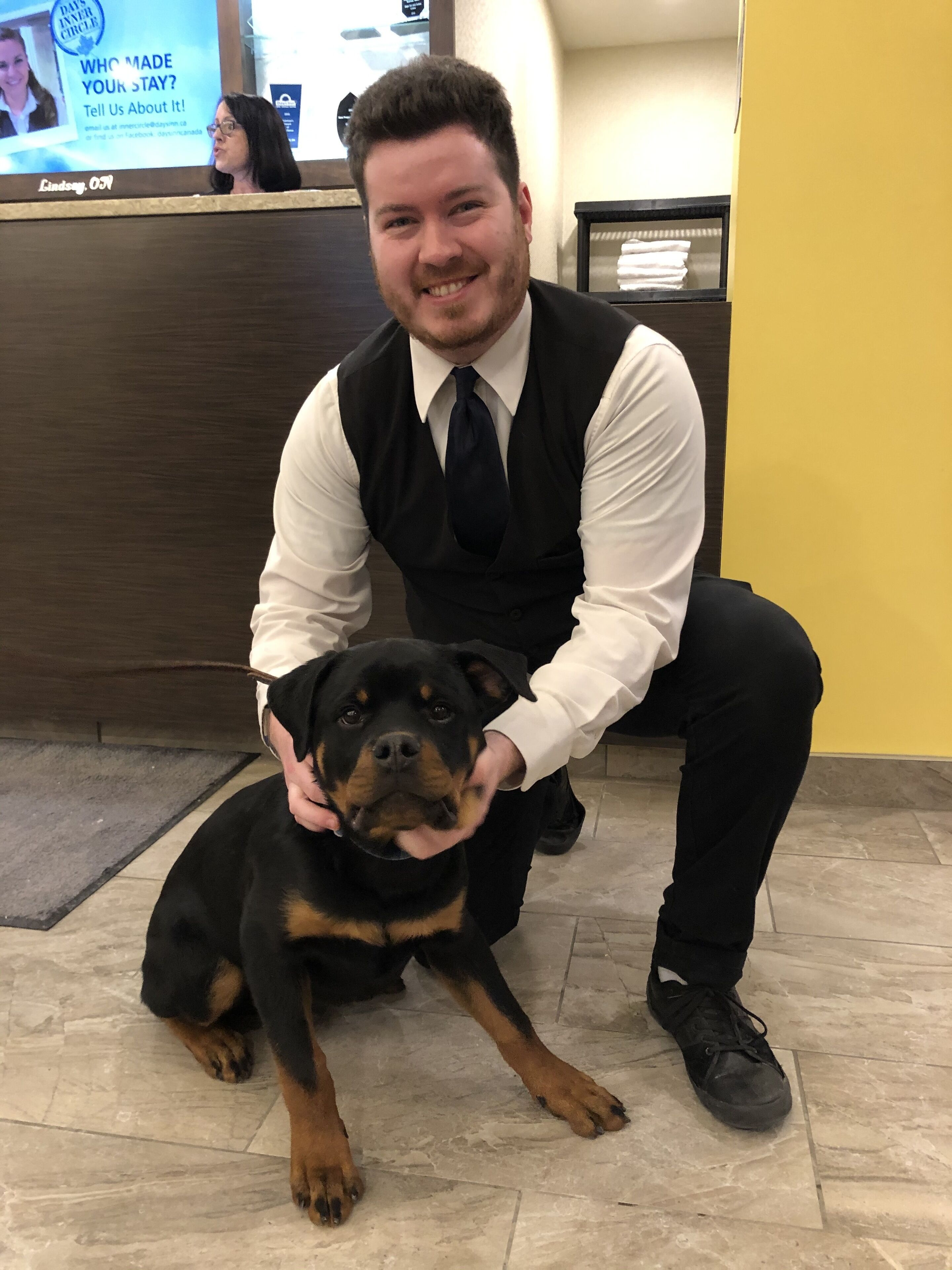 Uma pessoa sentada com um cachorro em uma área de recepção com balcão de madeira e tela digital ao fundo.