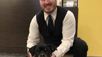 Uma pessoa sentada com um cachorro em uma área de recepção com balcão de madeira e tela digital ao fundo.