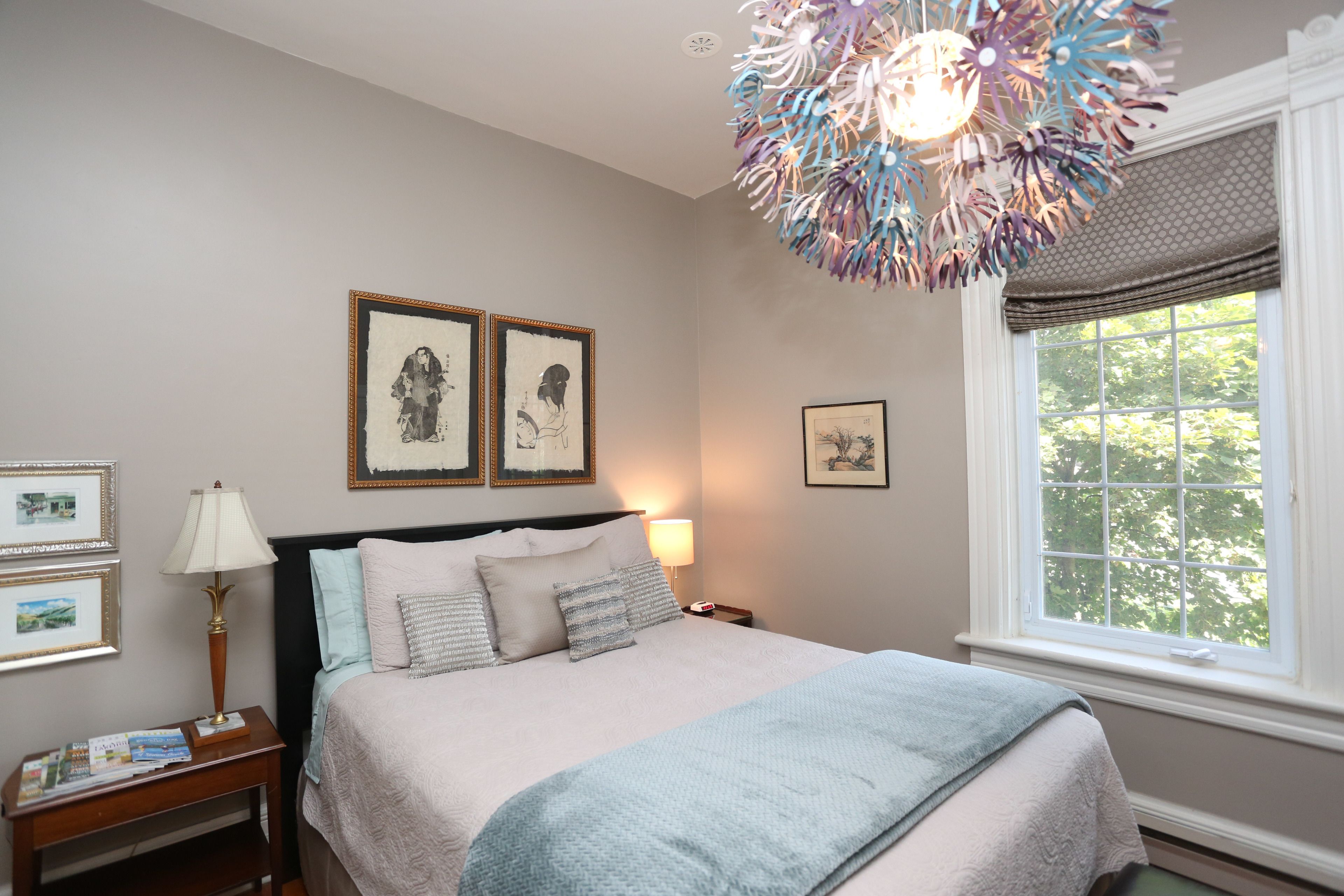Quarto com cama, mesas de cabeceira, quadros emoldurados, janela com vista e luminária decorativa suspensa.