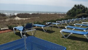 Uma área de descanso à beira-mar, com fileiras de espreguiçadeiras azuis e brancas e uma mesa azul, com vista para o oceano.