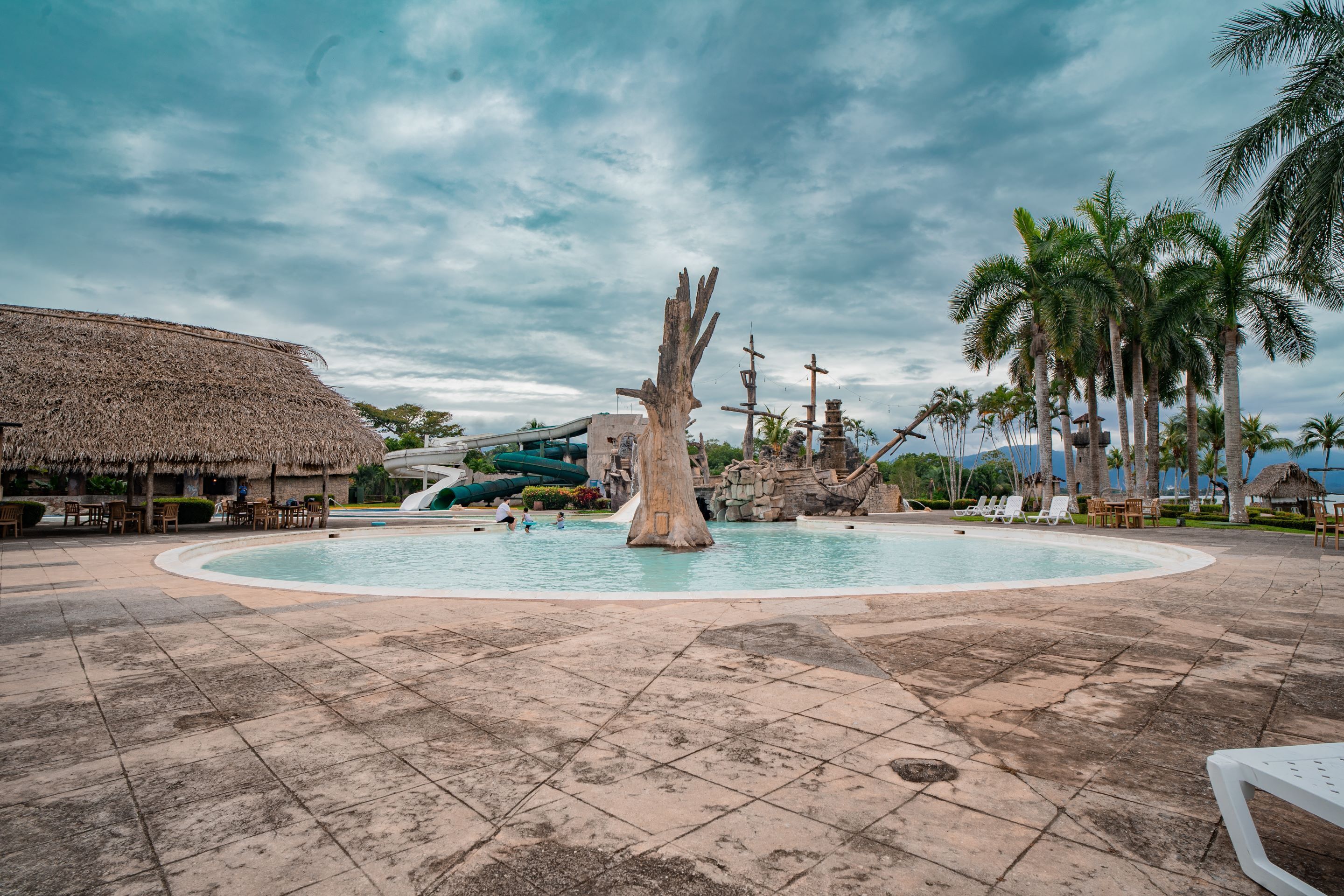 Área de piscina em resort com uma fonte central, palmeiras e um prédio com telhado de palha.