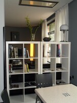 Quarto moderno com uma estante branca cheia de livros e objetos decorativos, uma pequena mesa com superfície branca e uma janela com cortinas brancas.