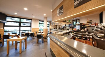 Um café moderno com mesas e cadeiras de madeira, um balcão com prateleiras cheias de garrafas e amplas janelas que permitem a entrada de luz natural.