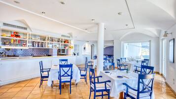 Restaurante com cadeiras azuis e mesas brancas, chão de azulejos e um balcão com diversos itens.