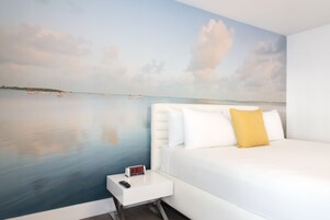 Um quarto com um mural de um mar tranquilo e barcos, uma cama branca com almofada amarela e uma mesa de cabeceira com relógio e controle remoto.