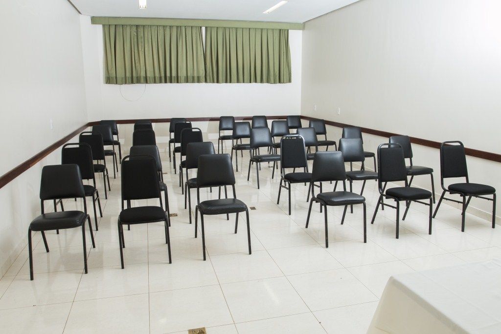 Uma sala de conferências com fileiras de cadeiras pretas, uma cortina verde e piso de azulejos brancos.