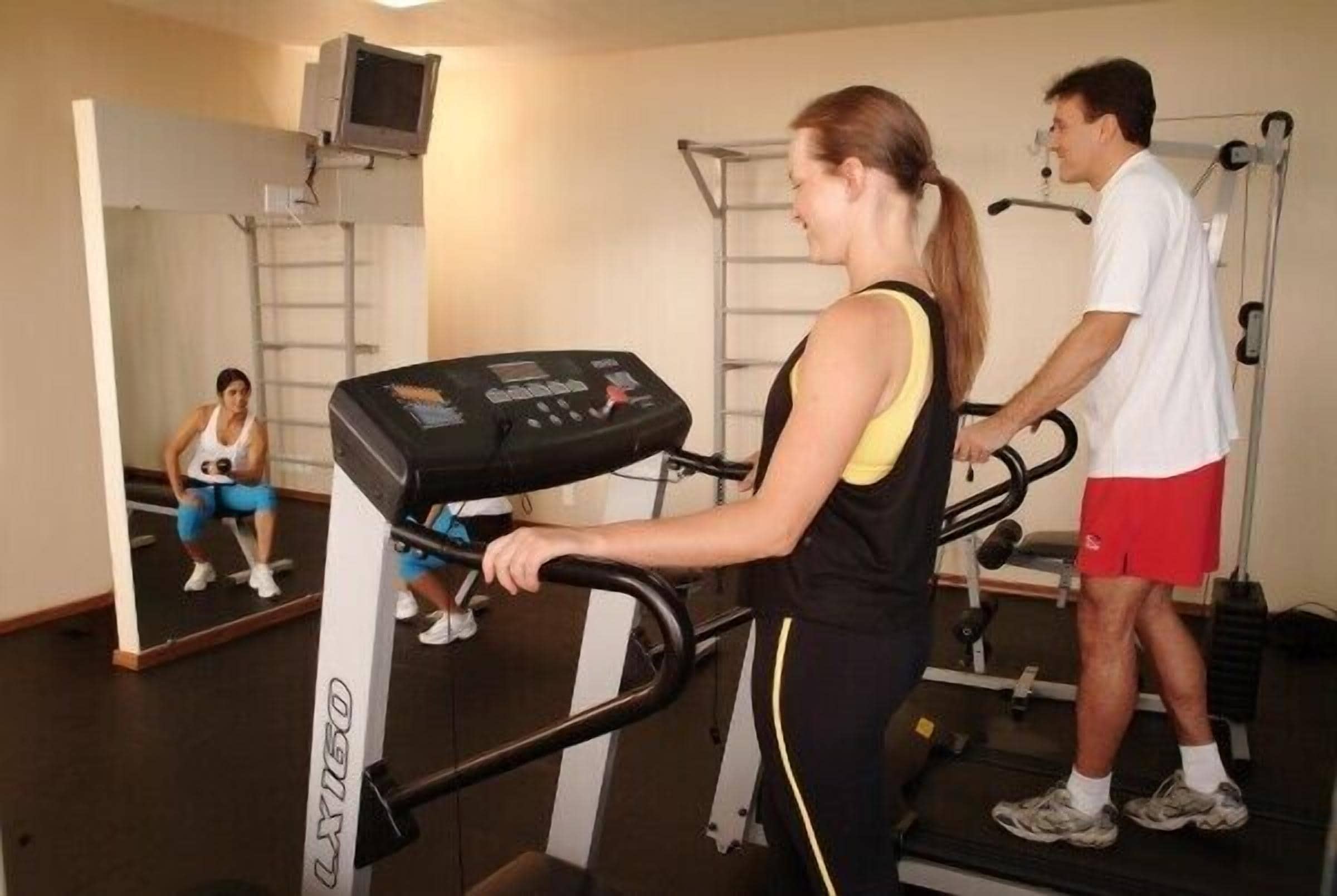 Ginásio com equipamentos de cardio, espelho e televisão na parede.