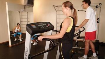 Ginásio com equipamentos de cardio, espelho e televisão na parede.