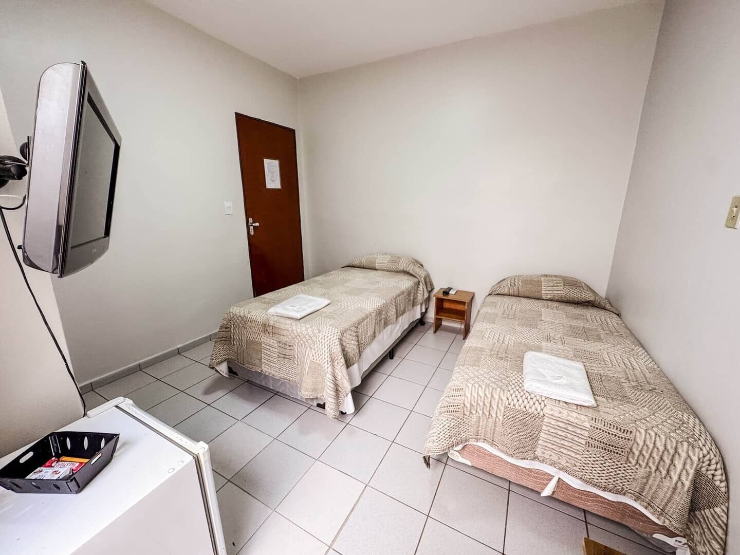 Quarto pequeno e limpo com duas camas de solteiro, porta de madeira, televisão na parede e uma pequena mesa com uma cesta.