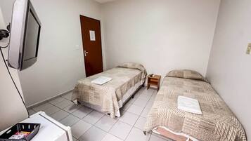 Quarto pequeno e limpo com duas camas de solteiro, porta de madeira, televisão na parede e uma pequena mesa com uma cesta.