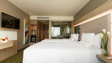 Quarto de hotel moderno com uma cama grande, uma TV fixada na parede, uma mesinha de cabeceira com um vaso de flores e vista para o banheiro.