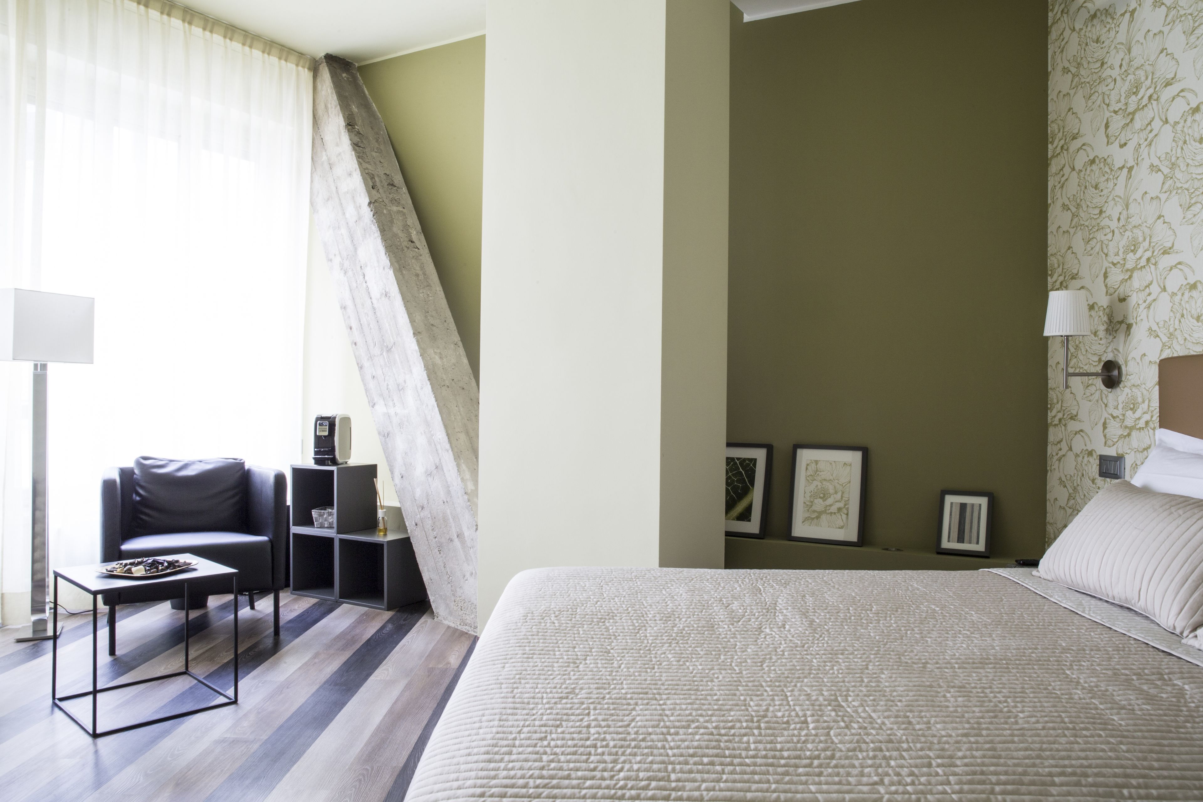 Quarto de hotel com uma cama grande, área de estar com uma poltrona e uma mesinha, mesa de cabeceira com abajur e janela com cortinas leves.