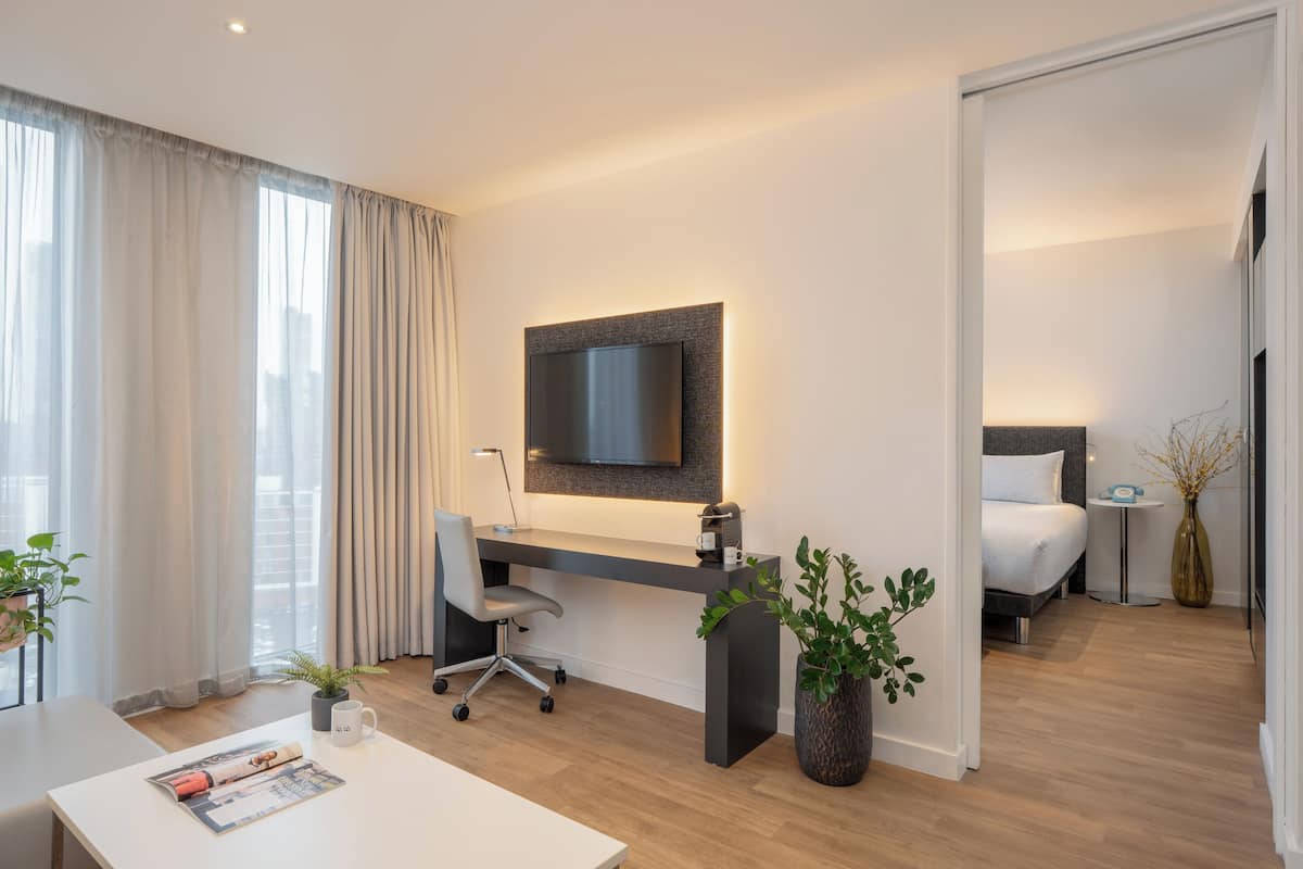 Quarto de hotel moderno com escrivaninha, cadeira, televisão, cama, mesa de cabeceira e uma planta em vaso.