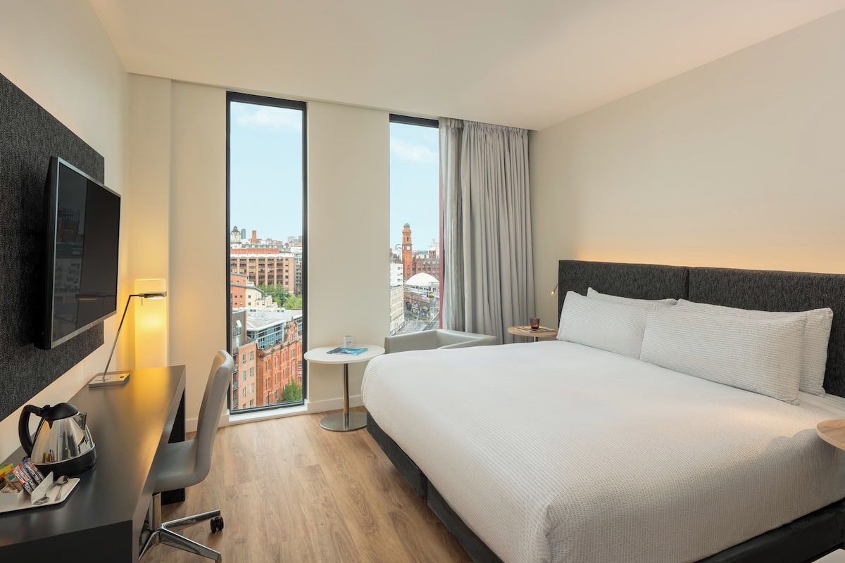 Quarto de hotel moderno com uma cama grande, escrivaninha, cadeira, TV e vista panorâmica para a cidade através de amplas janelas.